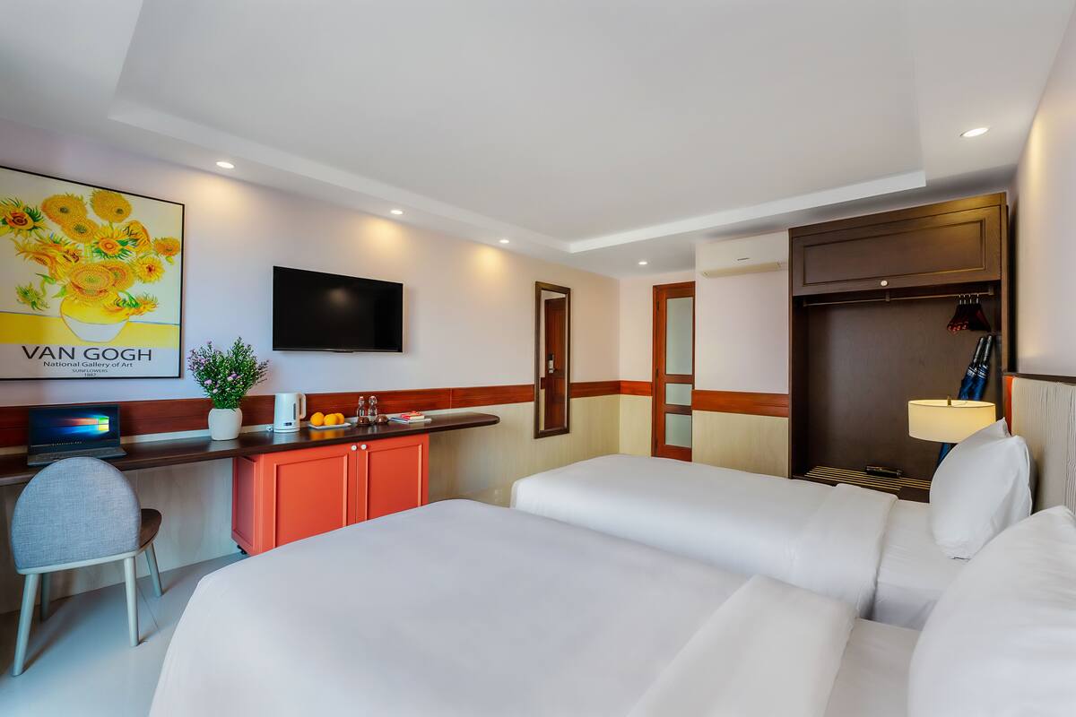Quarto de hotel com duas camas, uma escrivaninha com computador, televisão e um quadro de girassóis.