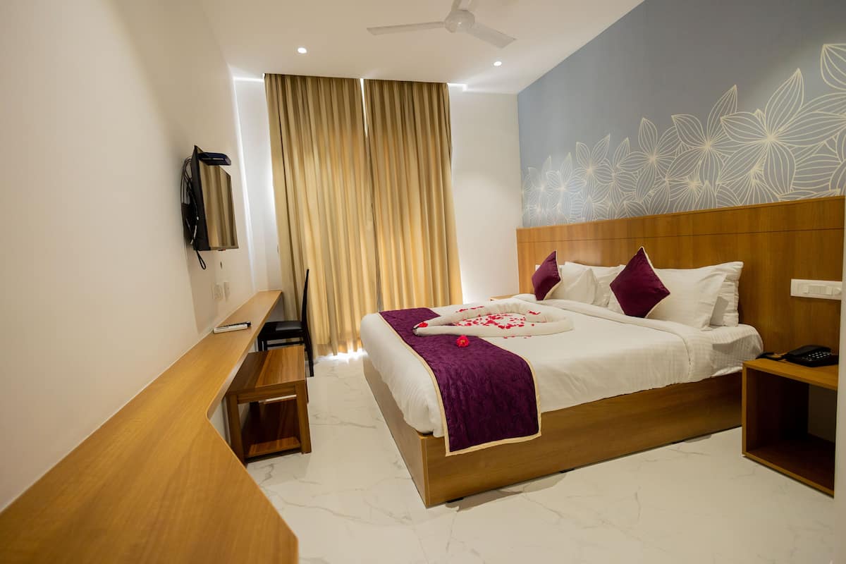 Quarto de hotel moderno com cabeceira de madeira, cama com roupa de cama branca e manta roxa, escrivaninha com cadeira e uma grande janela com cortinas.