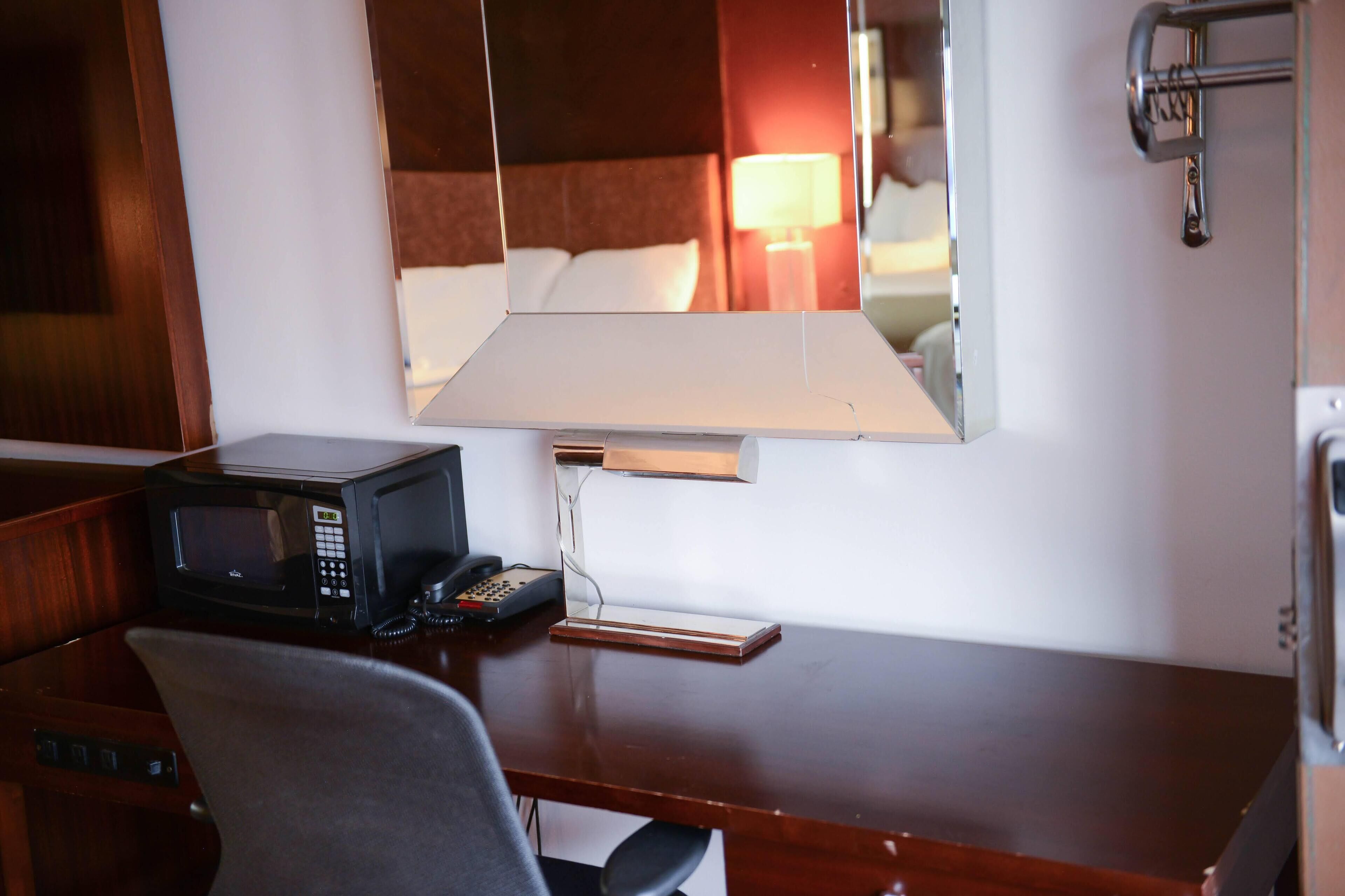 Quarto de hotel com uma escrivaninha de madeira, micro-ondas, telefone e luminária de cabeceira.