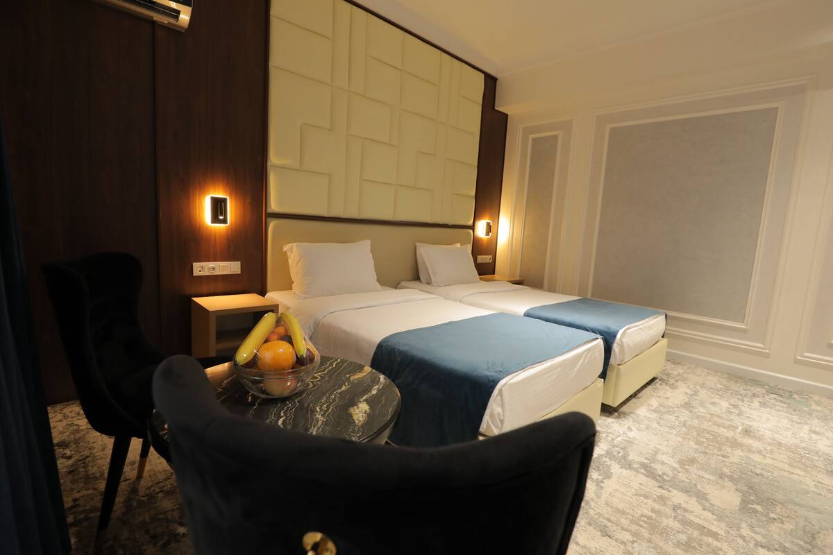 Quarto de hotel com duas camas, uma mesa pequena com frutas e uma cadeira.