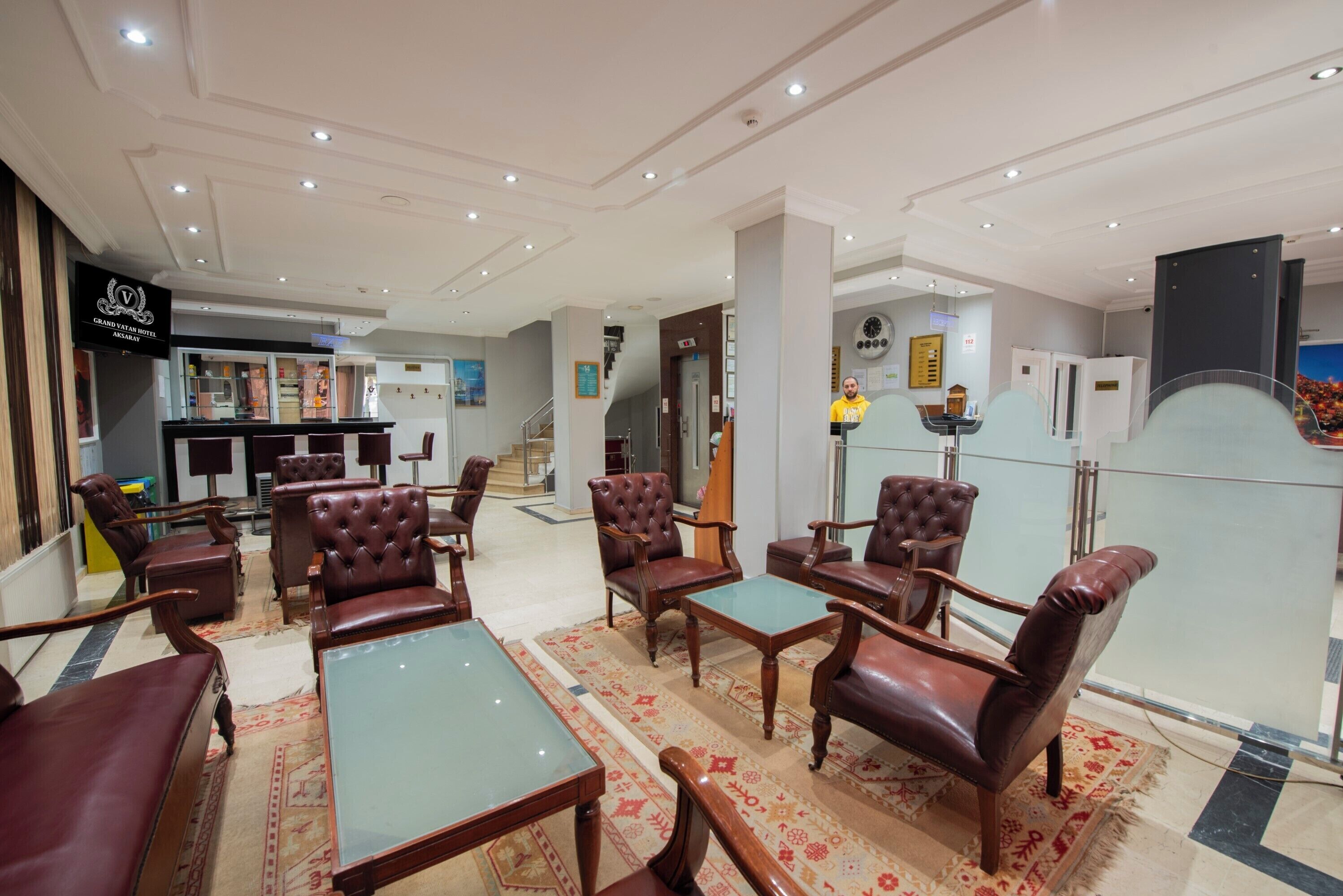 Um saguão de hotel com poltronas de couro, um balcão de recepção e uma escadaria que leva ao andar superior.
