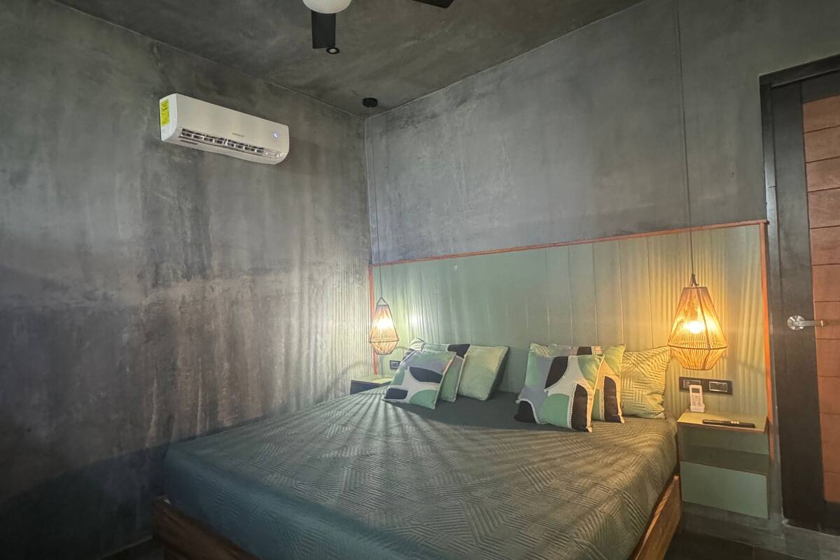 Quarto com cama, ventoinha no teto, aparelho de ar condicionado, abajures e porta de madeira.