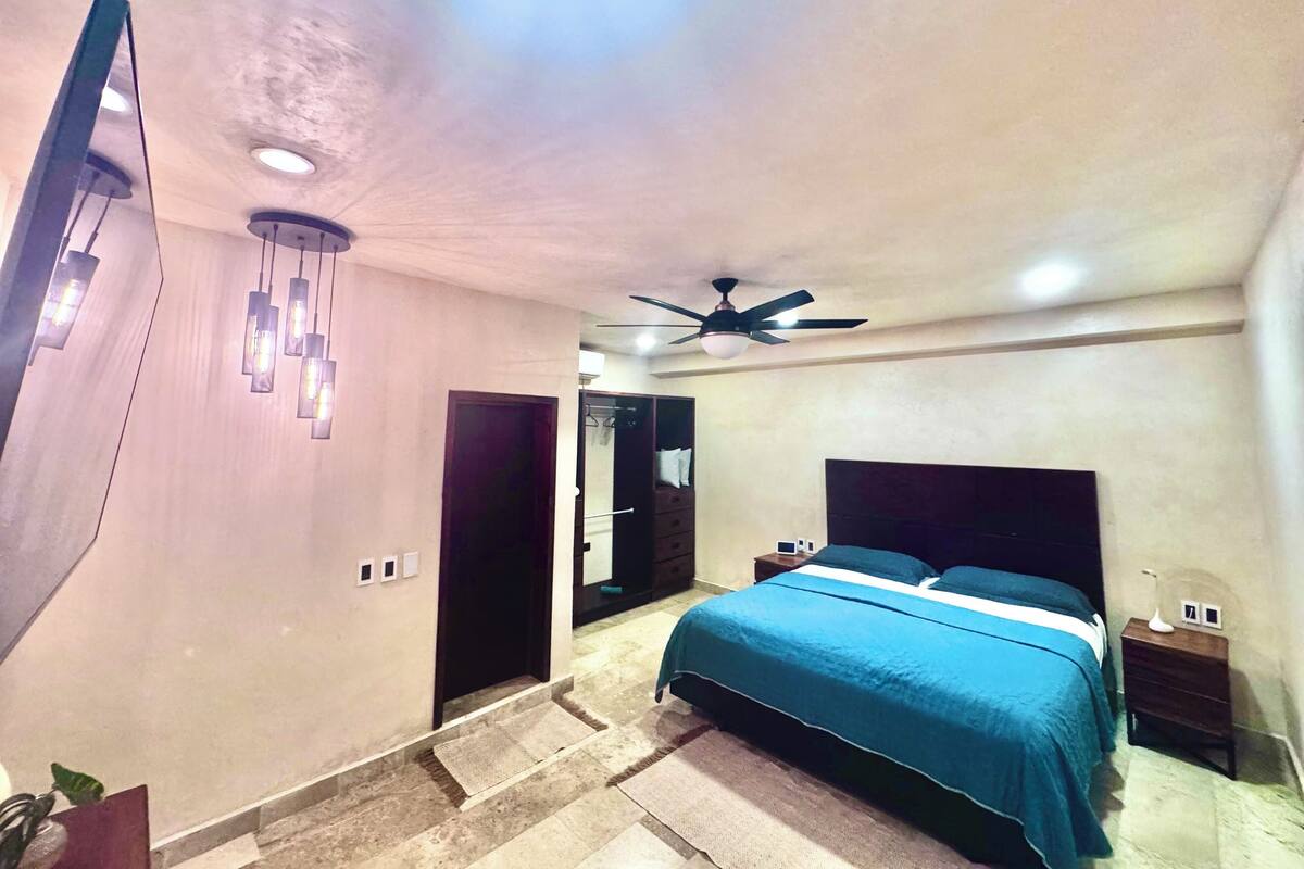 Quarto com cama, ventilador de teto, mesa de cabeceira e televisão fixada na parede.