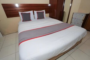 Uma cama bem arrumada com roupa de cama branca e detalhes vermelhos, uma cabeceira de madeira e piso de azulejo.