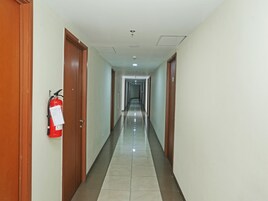 Um corredor comprido e estreito, com portas de madeira em ambos os lados, e um extintor de incêndio fixado na parede esquerda.