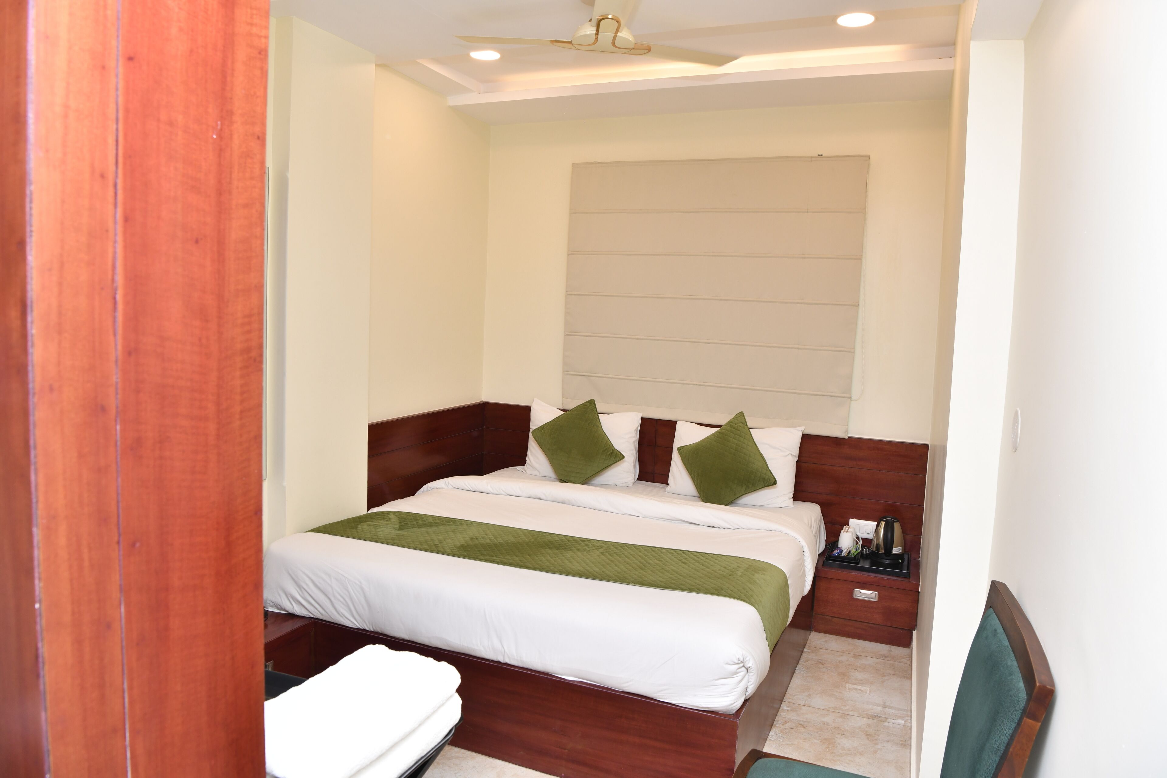 Quarto de hotel com cabeceira de madeira, cama com roupa de cama branca e almofadas verdes, mesa de cabeceira com abajur e criado-mudo com uma chávena e caixa de lenços.