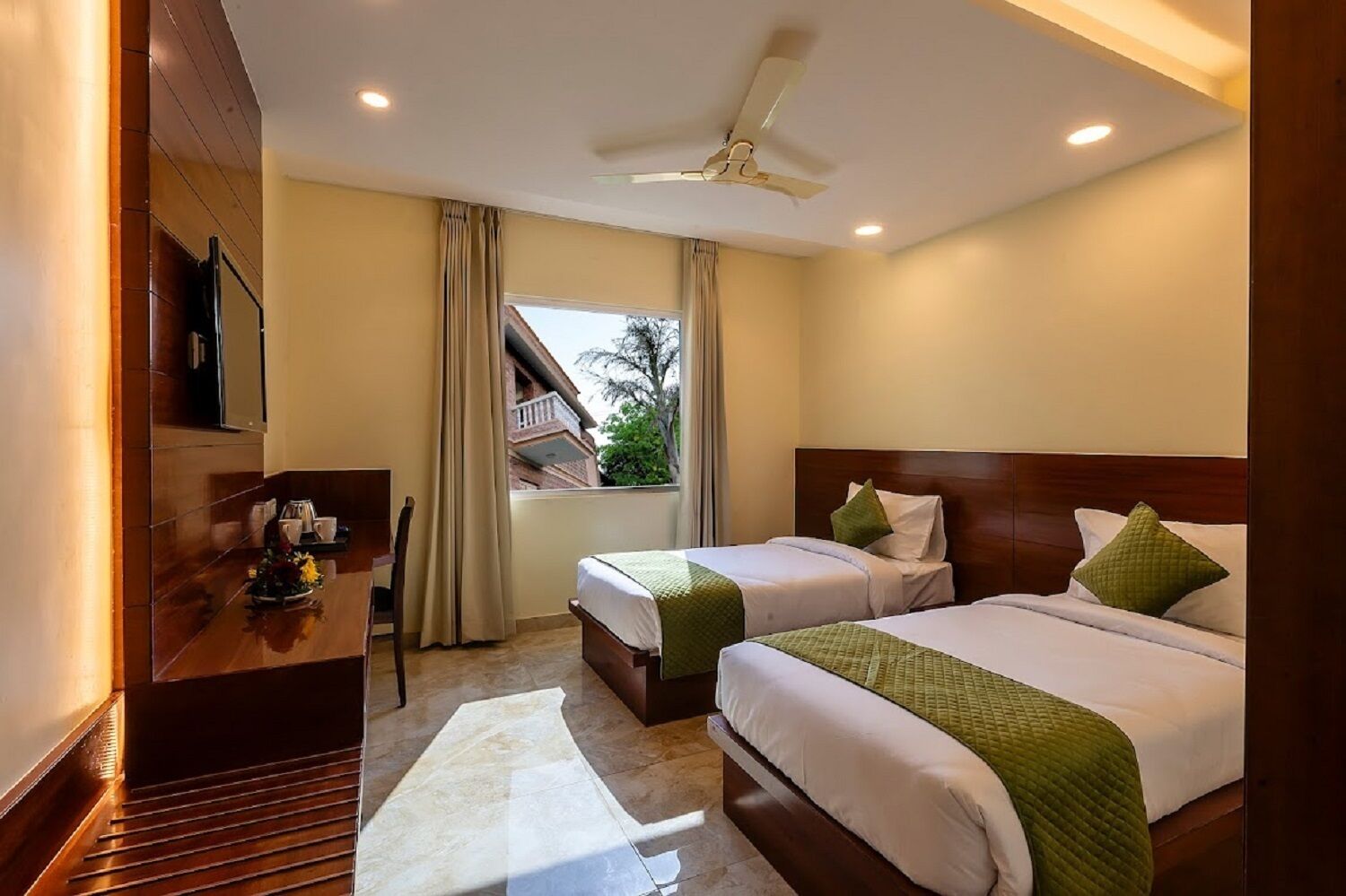 Quarto de hotel com duas camas, uma escrivaninha com cadeira, um ventilador de teto e uma janela com cortinas.