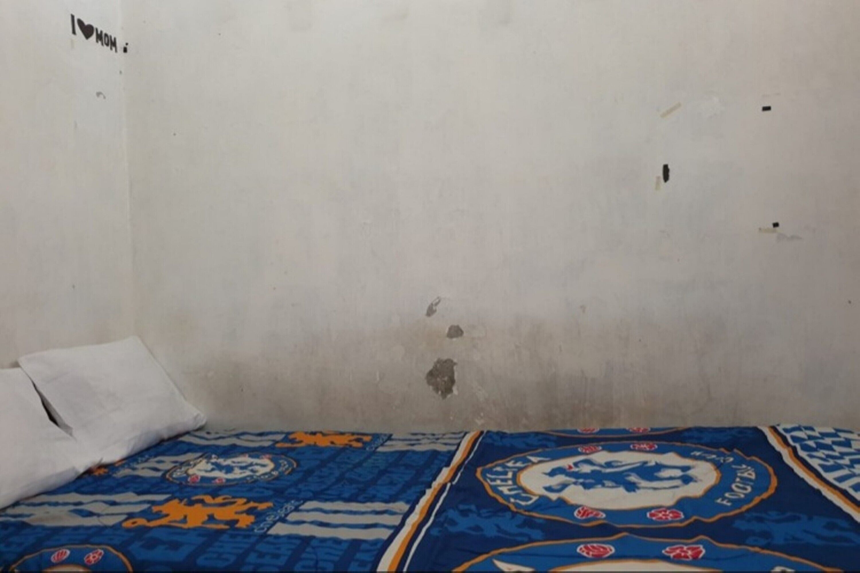 Quarto com cama coberta por um cobertor estampado, travesseiro branco e parede com algumas marcas e danos.