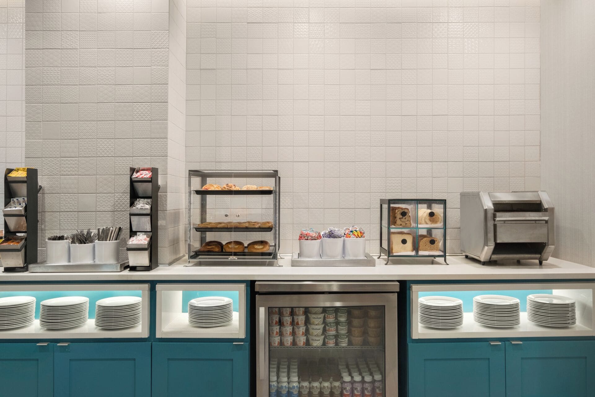Uma área de pequeno-almoço com serviço, incluindo um dispensador de bebidas refrigeradas, uma variedade de pães e uma máquina de café.