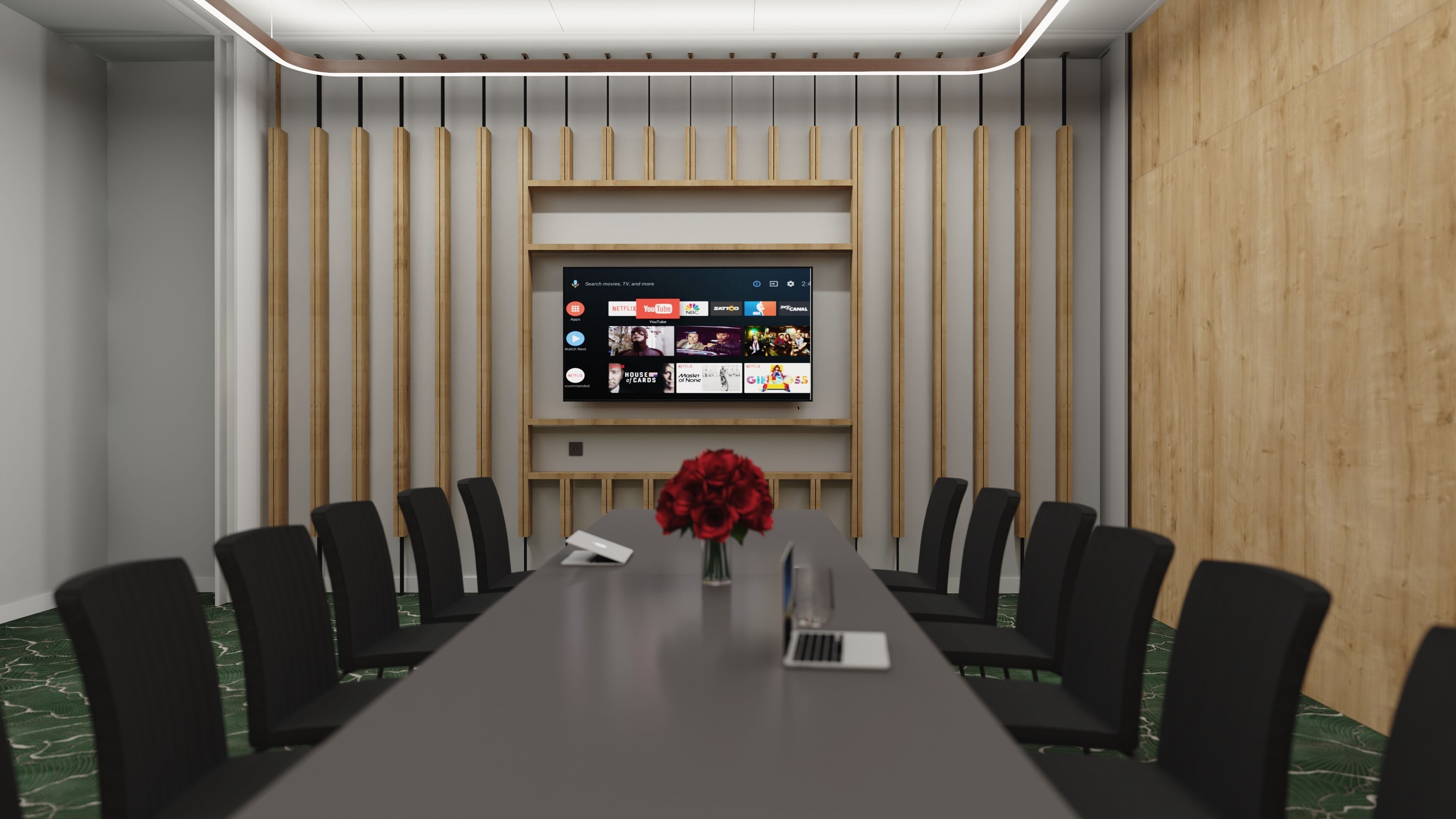 Uma sala de conferências com uma mesa comprida, cadeiras pretas, uma peça central florida e uma TV montada exibindo serviços de streaming.