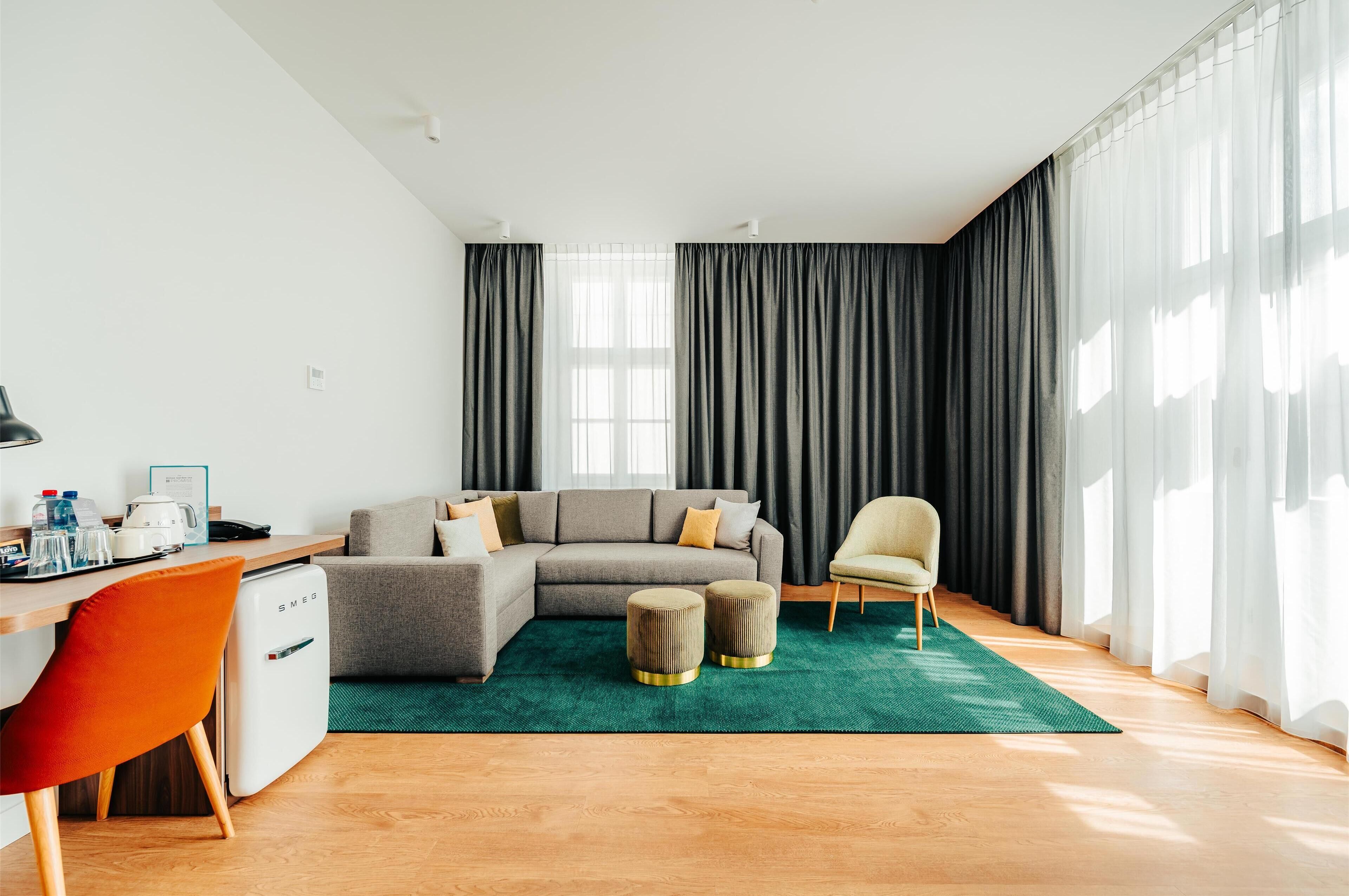 Uma sala de estar moderna com um sofá seccional cinza, uma mesa redonda pequena, uma cadeira vermelha, um mini-refrigerador branco e um tapete verde.