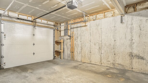 Uma garagem com porta branca, paredes de concreto e teto com vigas e fiação aparentes.