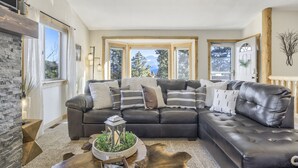 Uma sala de estar moderna com um sofá modular, lareira de pedra e grandes janelas com vista para árvores e montanhas.