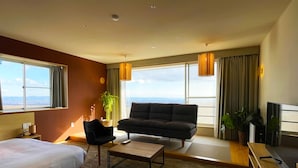 Quarto de hotel com cama, sofá, mesa de centro, televisão e uma grande janela com vista para a cidade.