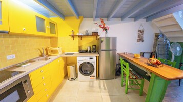 A kitchen with yellow cabinets, a washing machine, a refrigerator, and a dining area with a chair and table.