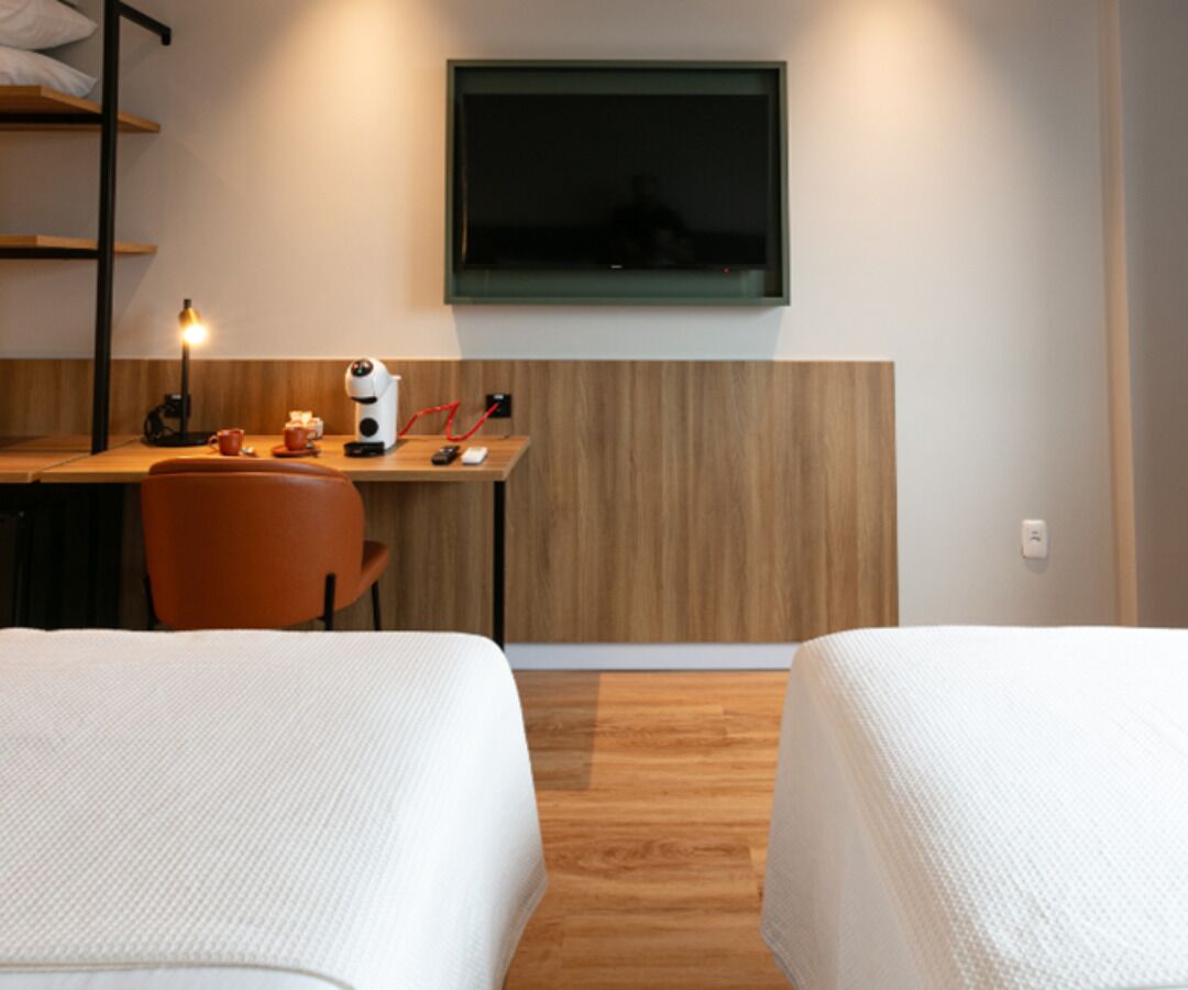 Quarto de hotel com duas camas, uma escrivaninha, uma cadeira, uma televisão na parede e um abajur.