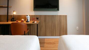 Quarto de hotel com duas camas, uma escrivaninha, uma cadeira, uma televisão na parede e um abajur.