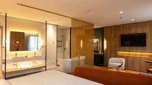 Quarto de hotel moderno com uma cama grande, área de estar com sofá e poltronas, chuveiro em box de vidro, TV de tela plana na parede e parede decorativa em madeira.