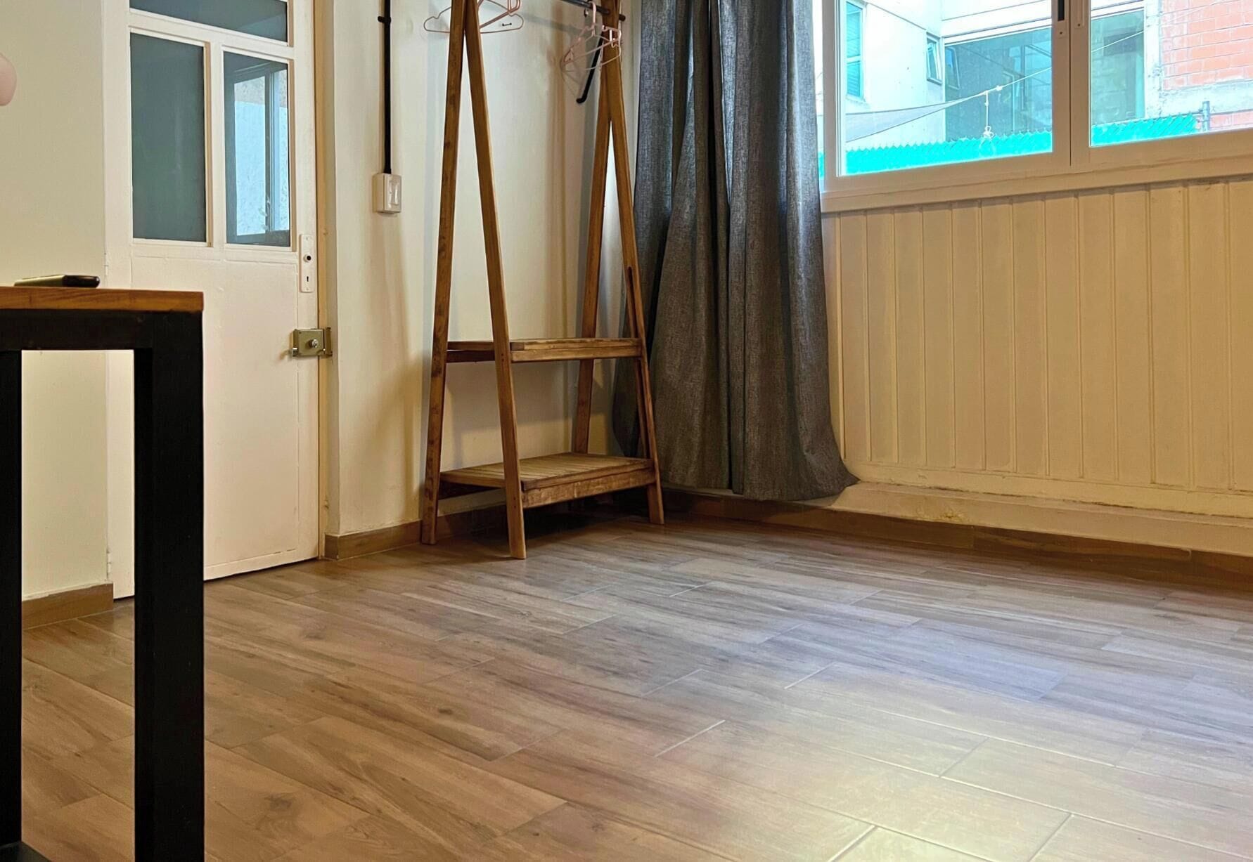 Um quarto com piso de madeira, uma porta, uma janela com cortinas e uma escada de madeira.