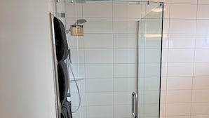 Casa de banho moderna com cabine de chuveiro em vidro, paredes com azulejos brancos e chão escuro.