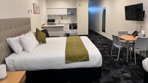 Quarto de hotel com cama, mesa de cabeceira e escrivaninha com cadeira. Cozinha compacta visível ao fundo.