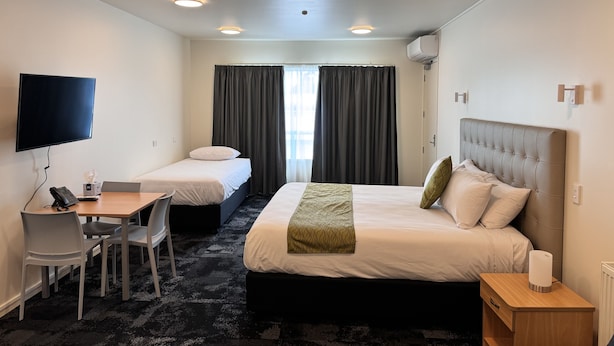 Quarto de hotel com cama, escrivaninha com cadeira, televisão e janela com cortinas.