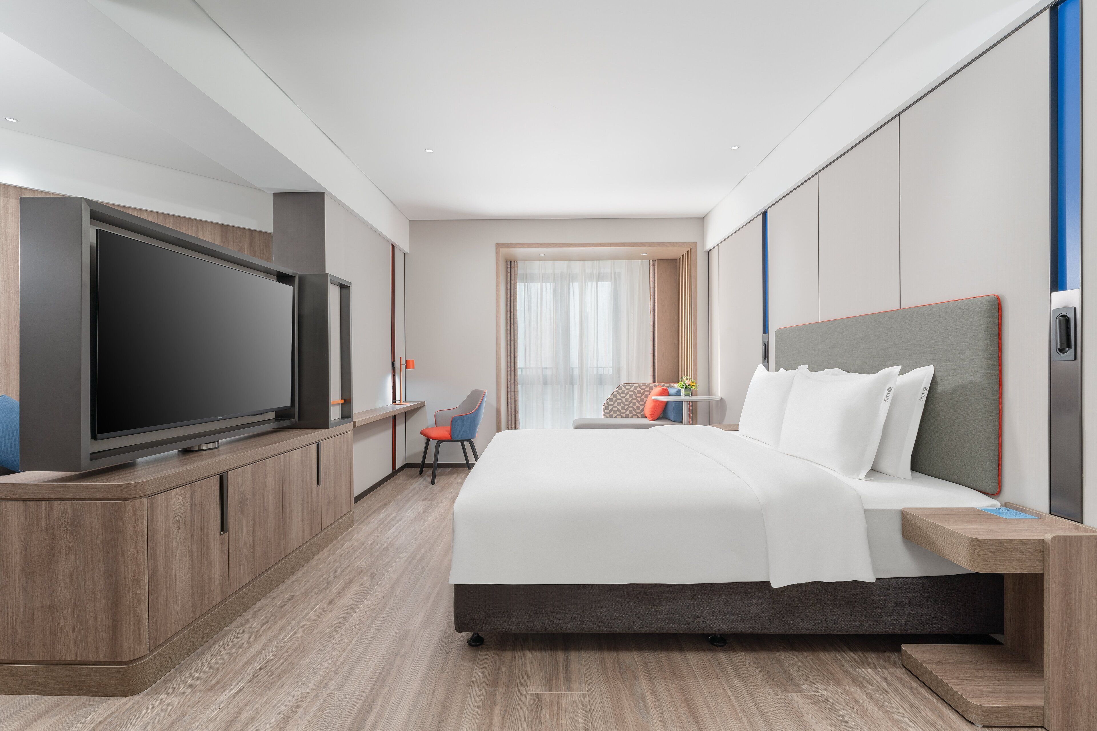 Quarto de hotel moderno com uma TV de tela grande, cama com roupa de cama branca, escrivaninha com cadeira e janela com cortinas finas.