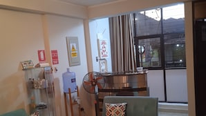 Uma sala de estar com dois sofás, uma mesa de centro de vidro e um tapete estampado. Há placas na parede, um ventilador e uma vista através de uma janela grande.