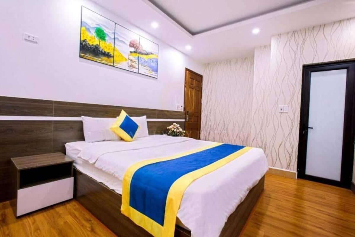 Quarto de hotel com uma cama, mesa de cabeceira, piso de madeira, porta e parede com papel de parede estampado.
