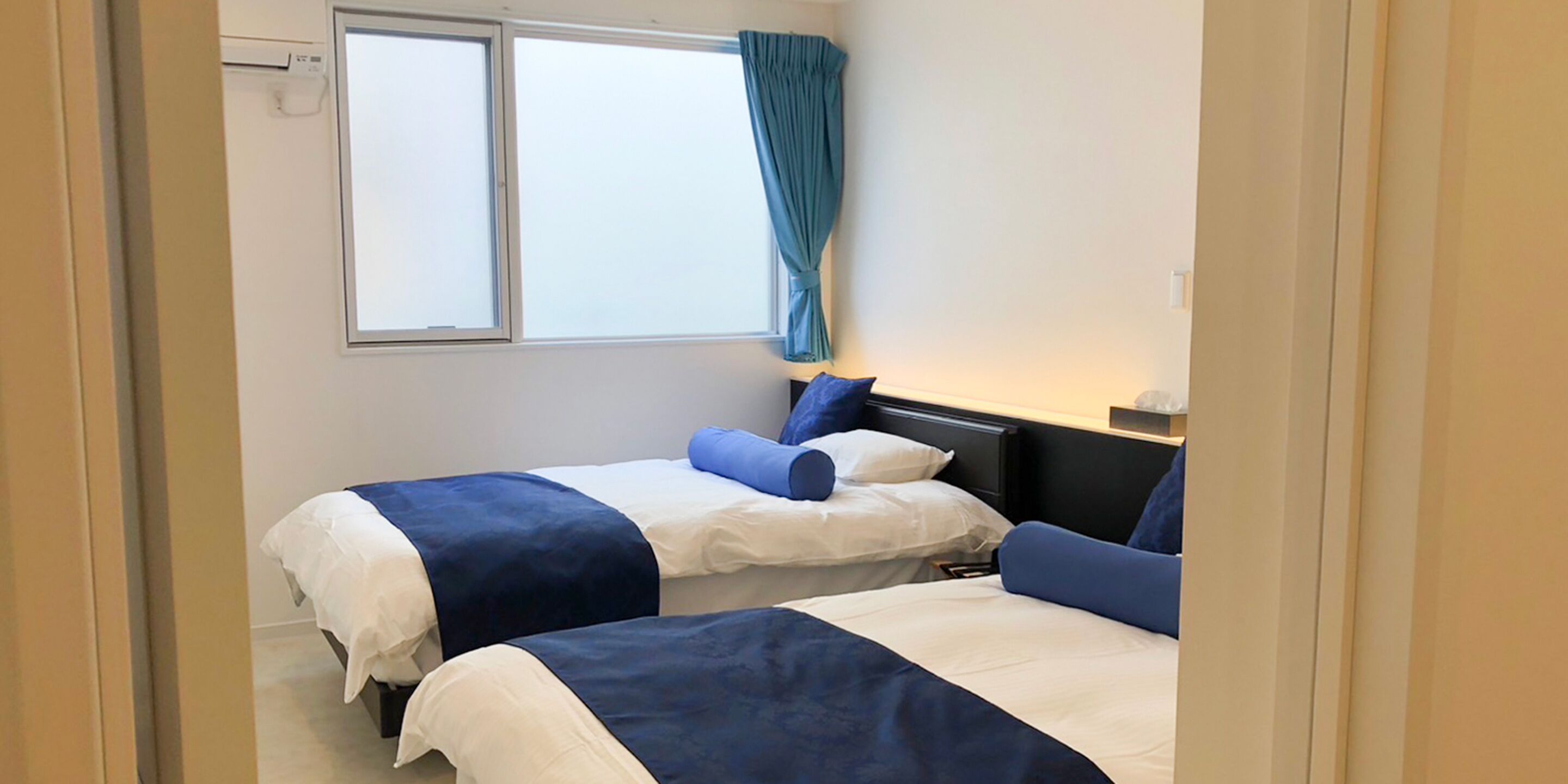 Quarto de hotel com duas camas, uma janela com cortinas e uma mesinha de cabeceira.