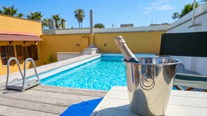 Área da piscina com um balde de metal contendo uma garrafa, um deck de madeira e uma piscina azul cristalina.