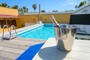 Área da piscina com um balde de metal contendo uma garrafa, um deck de madeira e uma piscina azul cristalina.