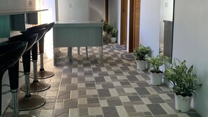 Um corredor com piso quadriculado, uma mesa de jantar com cadeiras e plantas em vasos.