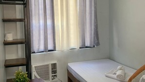 Quarto pequeno com uma cama de solteiro e beliche de madeira, uma mesinha com uma planta, uma janela com cortinas e um radiador.