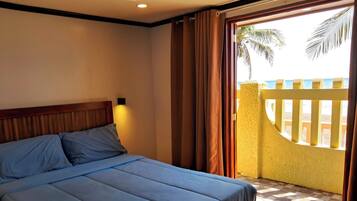 Quarto de hotel com cama, cortinas, luz acoplada à parede e vista para a praia.