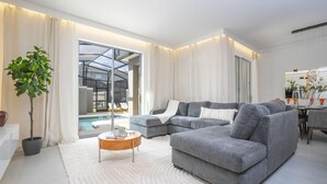 Uma sala de estar moderna com um sofá seccional cinza, uma mesa de centro redonda e uma grande janela com vista para uma piscina.