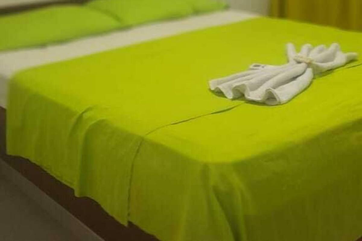 Cama com colcha verde, toalhas brancas, cabeceira listrada e cortinas amarelas.