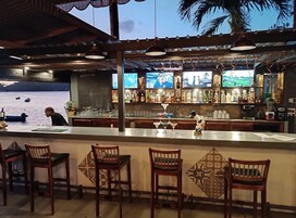 Zona de bar com vista para o mar, banquetas e balcão com garrafas e copos.