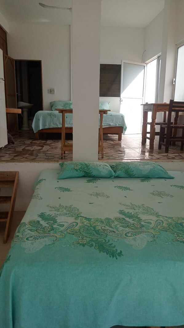 Quarto com cama, cadeira de madeira, mesinha e banheiro visível ao fundo.