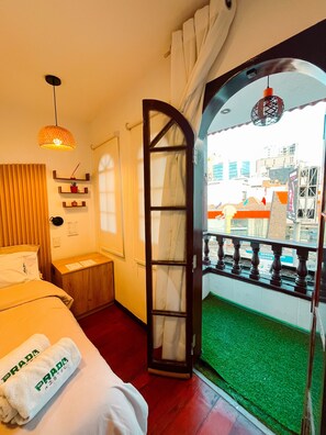 Quarto de hotel com cama, uma pequena mesa de cabeceira de madeira, prateleira com uma planta em vaso, luminária pendente e varanda com vista para a cidade.