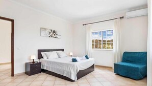 Quarto com cama, mesas de cabeceira, janela e uma cadeira azul.