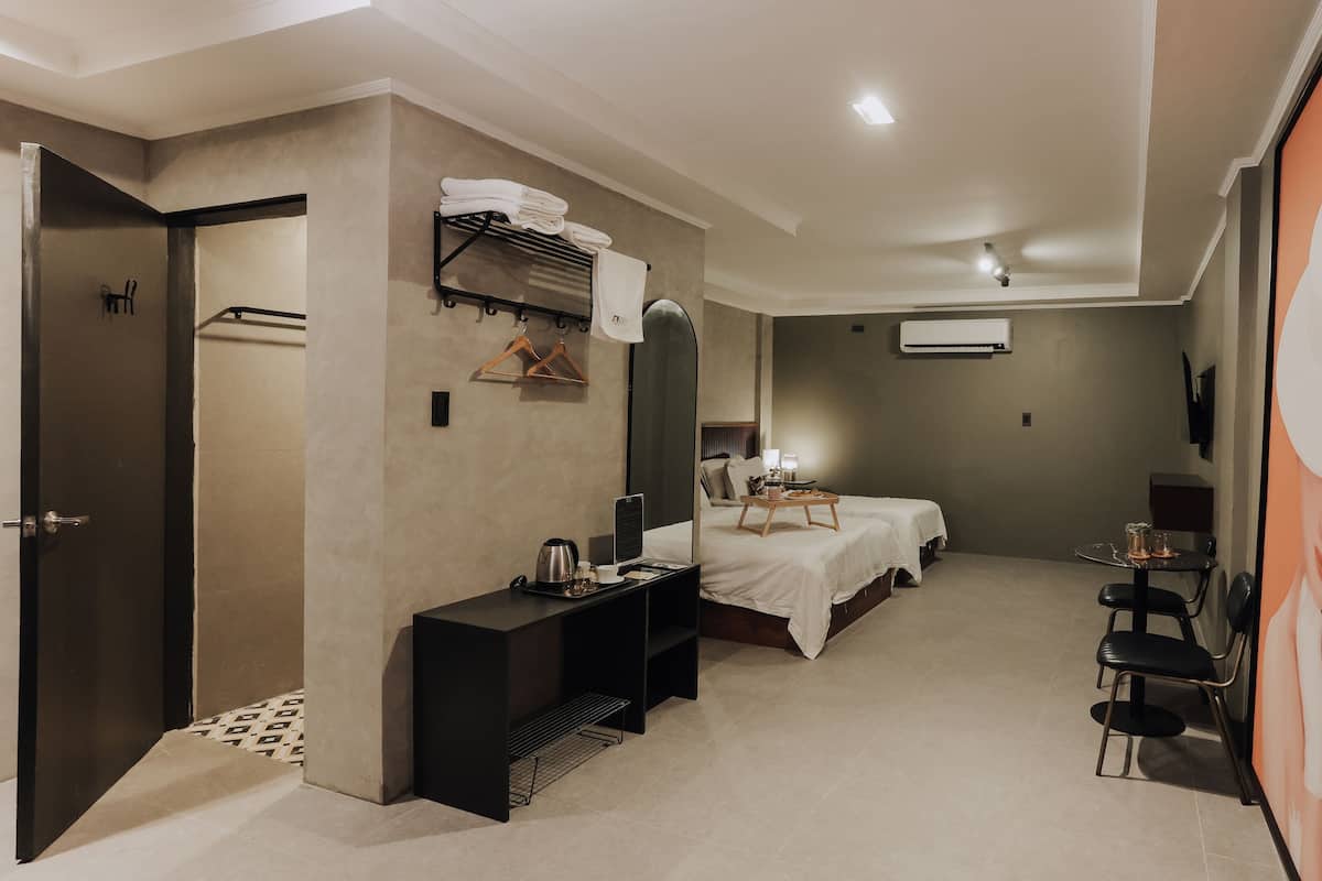 Quarto de hotel com cama, mesa de cabeceira e espelho. Há uma escrivaninha com cadeira e uma pequena mesa com abajur.