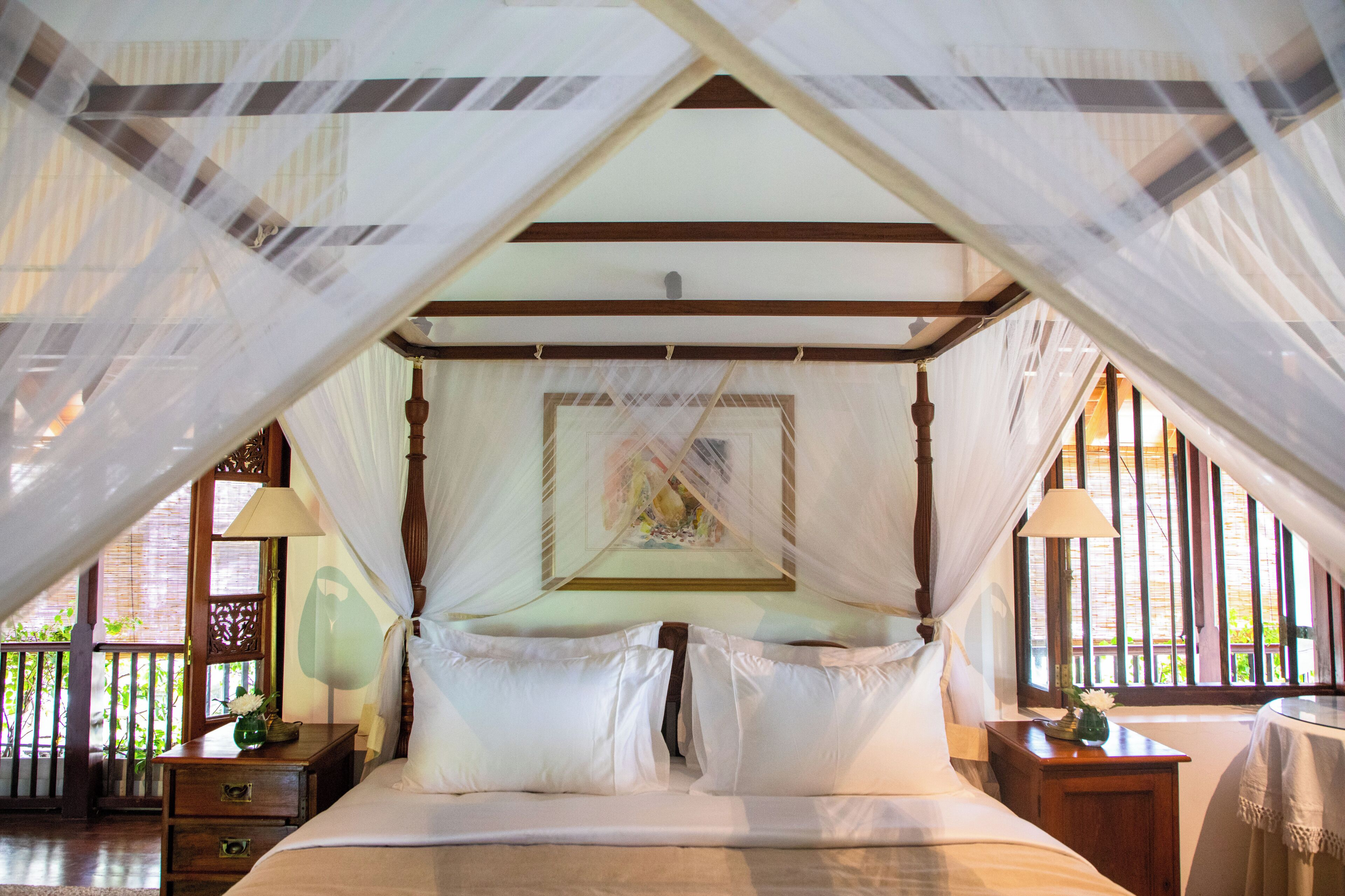 Uma cama de dossel com roupa de cama branca, cabeceira de madeira e um toldo. Há duas mesas de cabeceira com luminárias e um pequeno vaso sobre uma delas.
