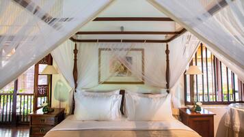 Uma cama de dossel com roupa de cama branca, cabeceira de madeira e um toldo. Há duas mesas de cabeceira com luminárias e um pequeno vaso sobre uma delas.