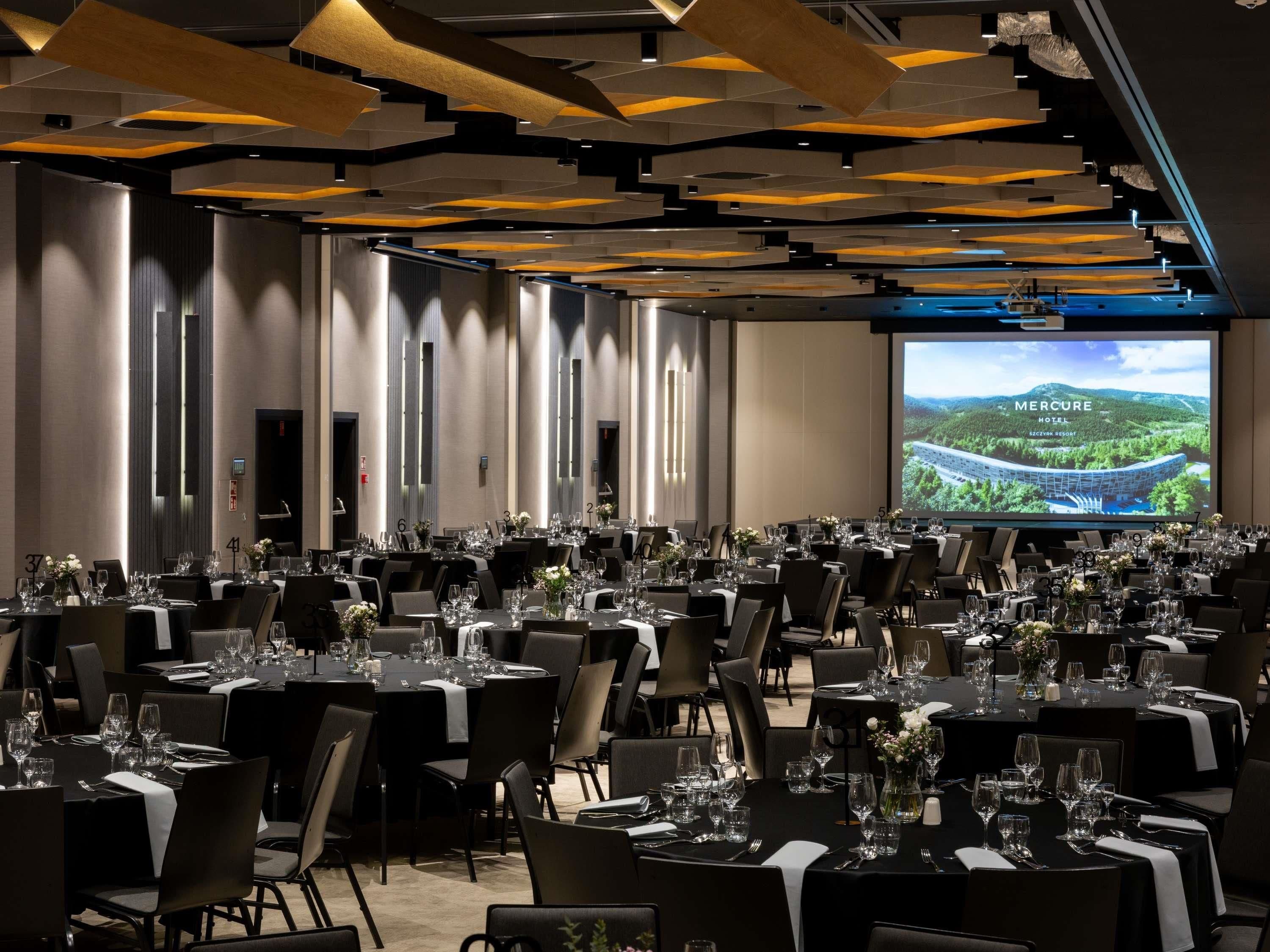 Uma sala de conferência com várias mesas preparadas para refeições, uma tela grande exibindo a imagem de um hotel e um design moderno no teto.