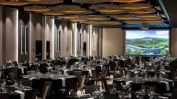 Uma sala de conferência com várias mesas preparadas para refeições, uma tela grande exibindo a imagem de um hotel e um design moderno no teto.