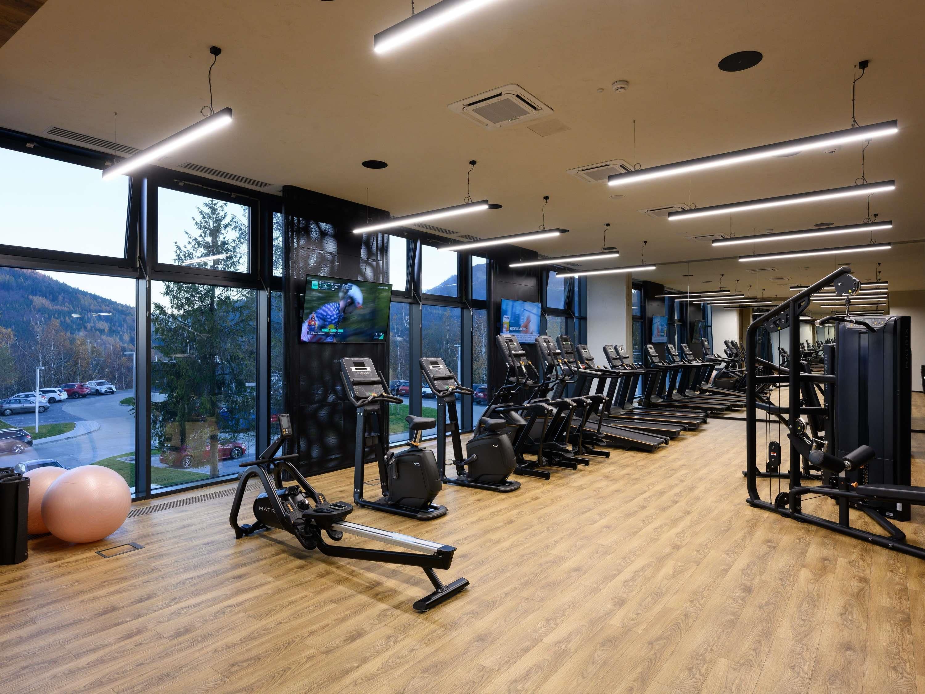 Academia com máquinas de cardio, estações de musculação e grandes janelas com vista para um estacionamento e árvores.