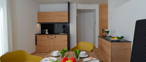 Uma área de refeições moderna com uma mesa de madeira posta para dois, cadeiras amarelas e uma pequena cozinha ao fundo.