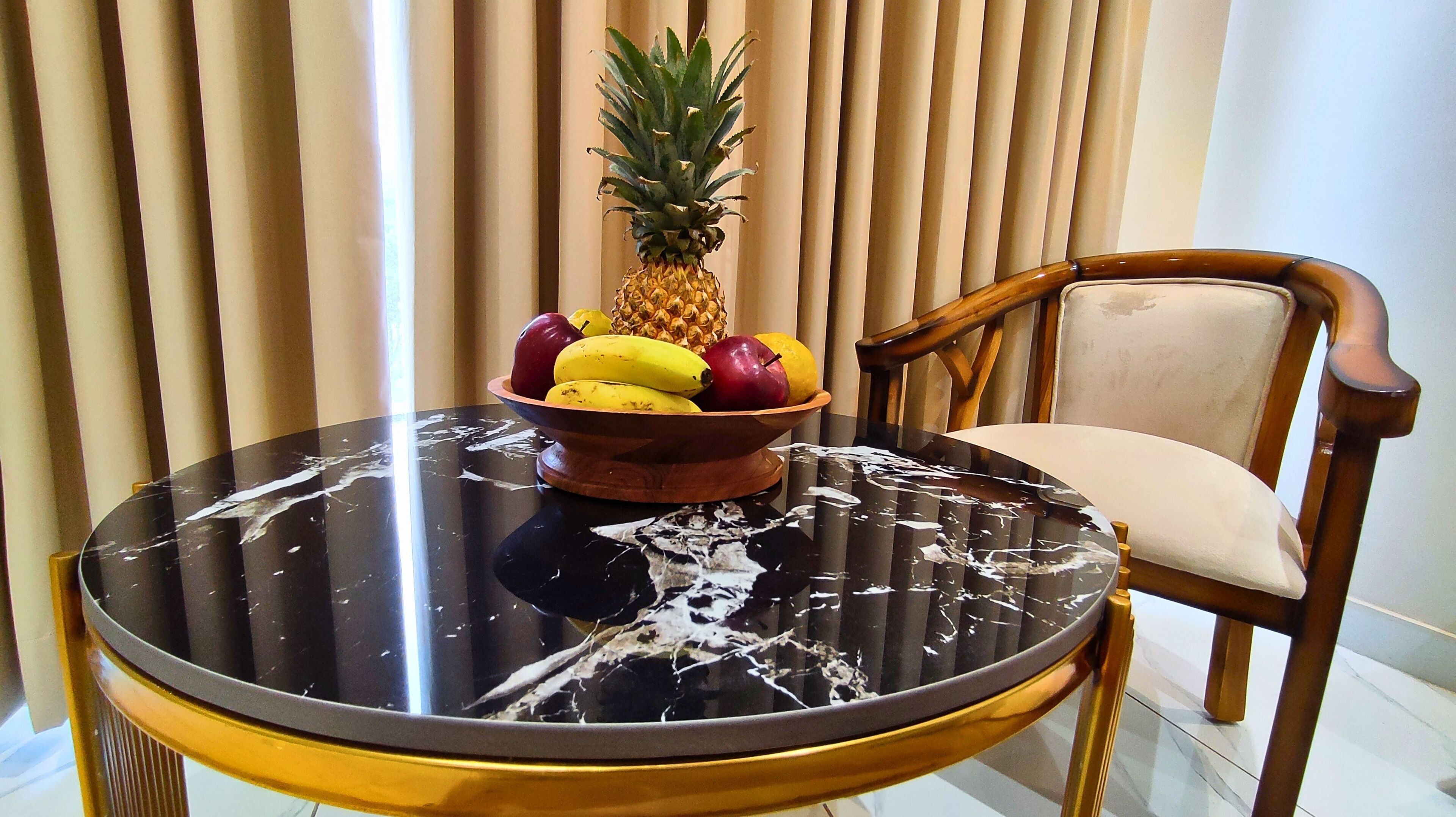 Uma mesa redonda com tampo de mármore preto, uma tigela de madeira com bananas, maçãs e uma abacaxi, e uma cadeira de madeira com assento acolchoado.
