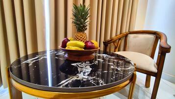 Uma mesa redonda com tampo de mármore preto, uma tigela de madeira com bananas, maçãs e uma abacaxi, e uma cadeira de madeira com assento acolchoado.